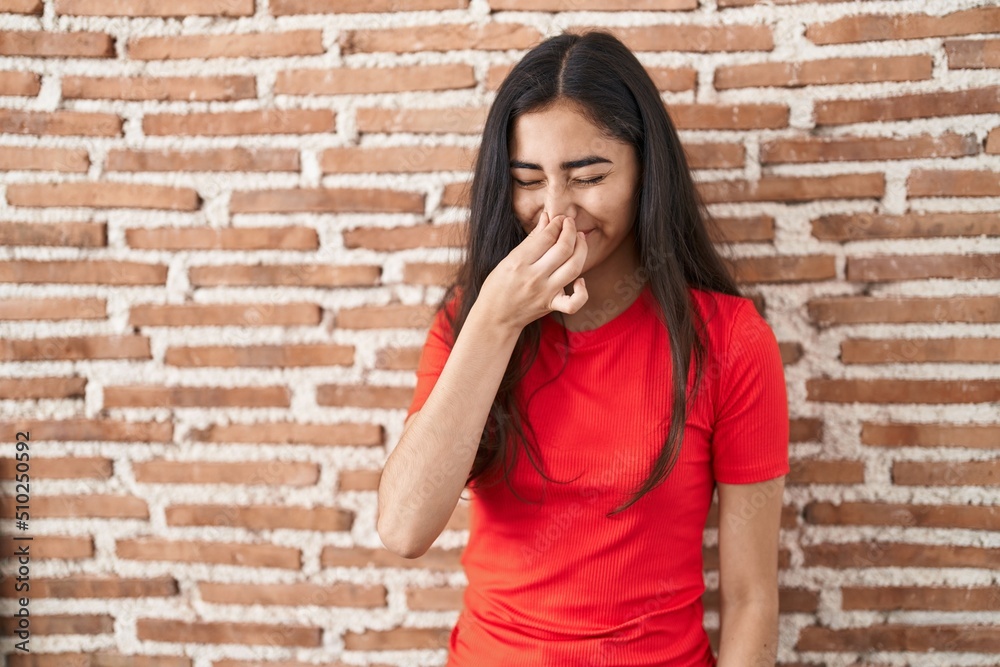 Young teenager girl standing over bricks wall smelling something stinky ...