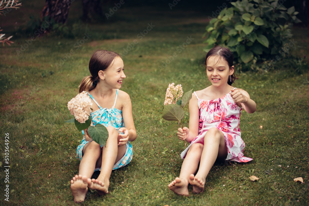 Two little barefoot girls in light summer dresses pose sitting on the ground in a summer park ...
