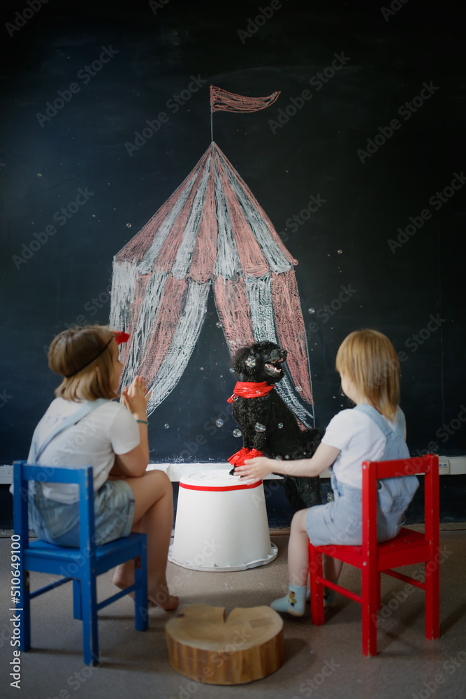 Two children watch poodle dog perform in front of painted circus ...