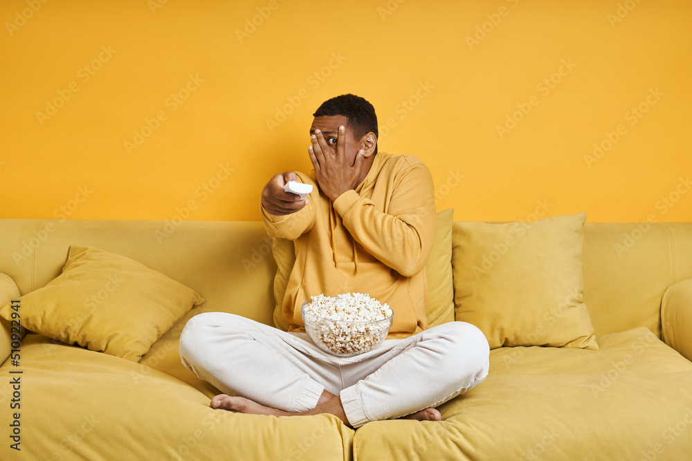 Young man watching TV and looking terrified while sitting on the couch ...