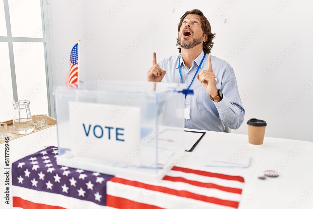 Handsome middle age man sitting at voting stand amazed and surprised ...