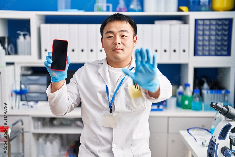 Young chinese man working at scientist laboratory holding smartphone ...