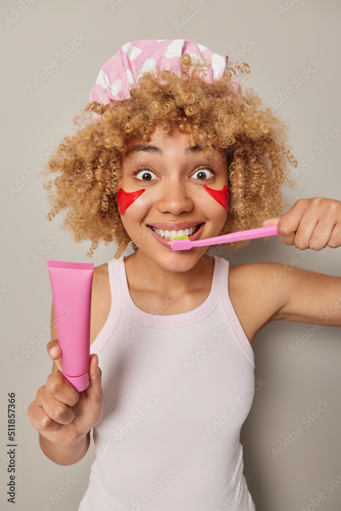 Cheerful woman with curly hair cleans teeth with toothbrush holds tube ...