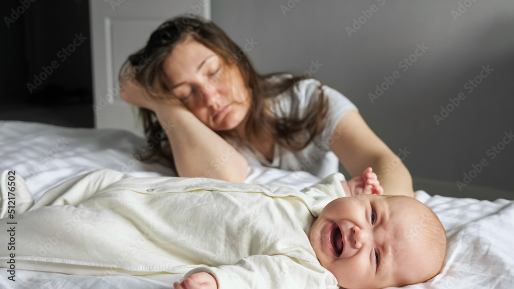 Tired mother sleeps sitting on floor leaning on bed with crying baby ...