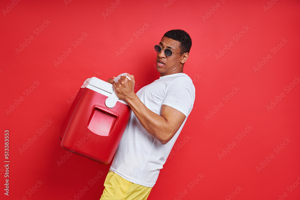 Young African man carrying heavy cooler box while standing against red ...