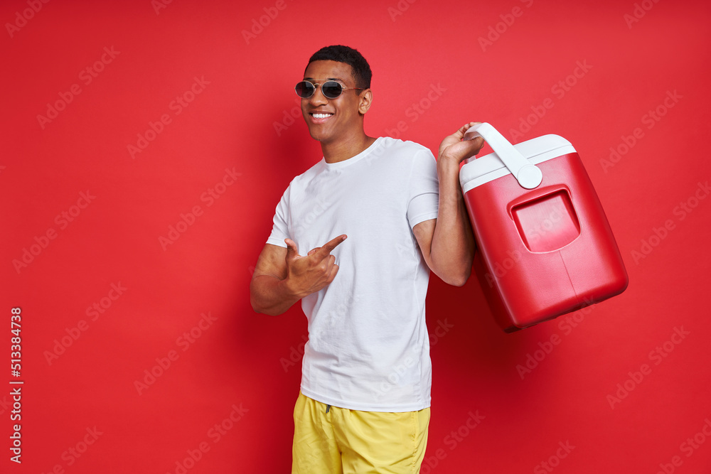 Cheerful African man pointing cooler box while standing against red ...
