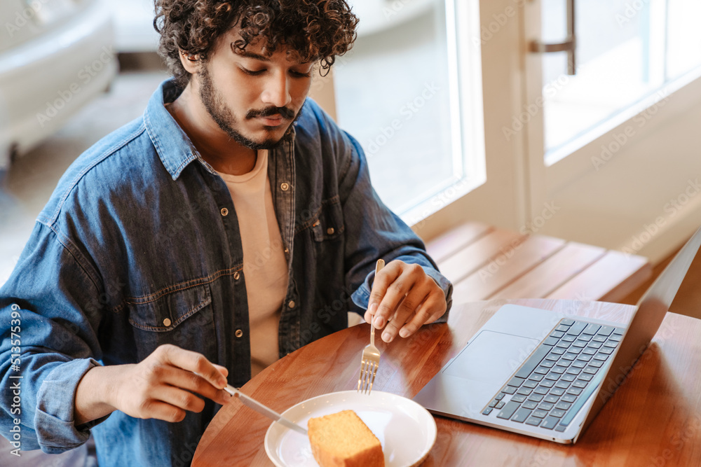 Young calm handsome indian curly man eating cake while working Stock ...