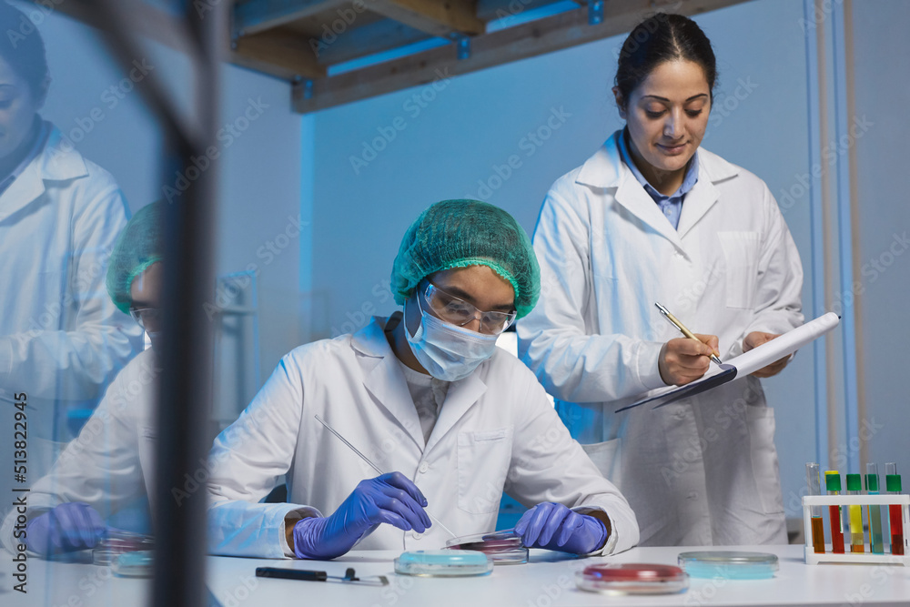 Young Indian assistant in lab coat making notes about changes of ...