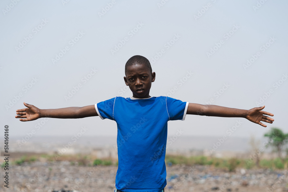 Worried African boy with outstretched arms standing in an arid field ...