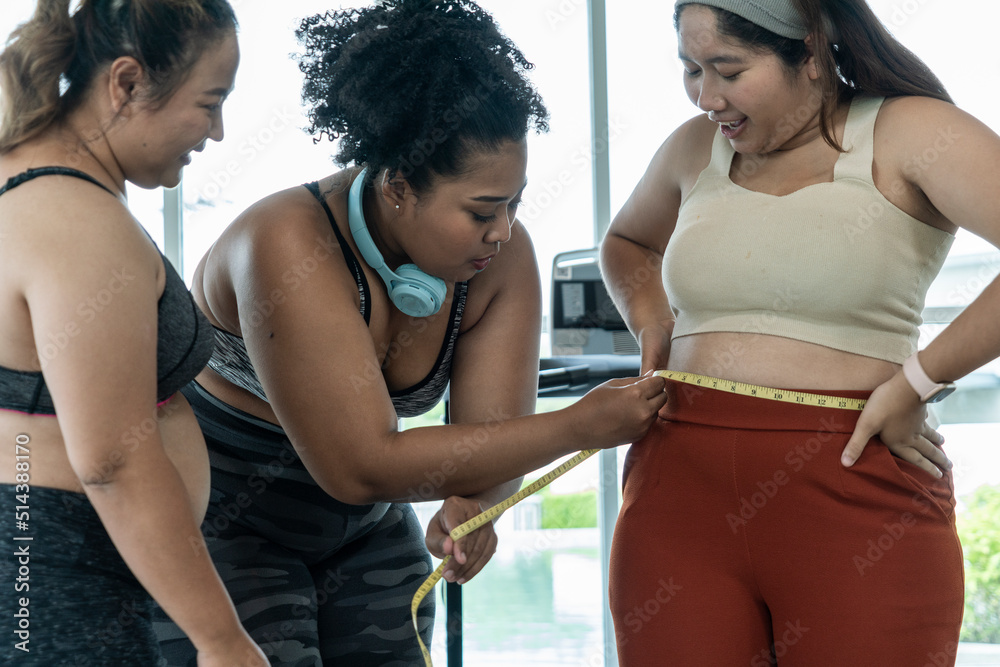 Three obese women in sports bras and overweight friends use a tape measure to measure their ...