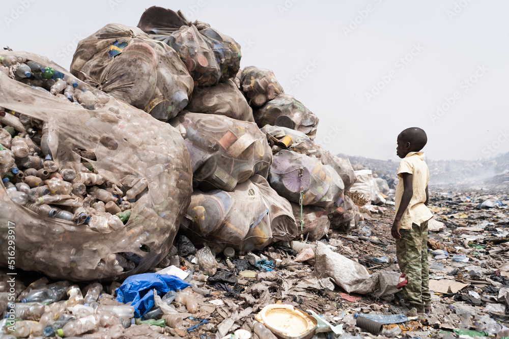 African child in a smoky landfill looking at giant bags with plastic ...