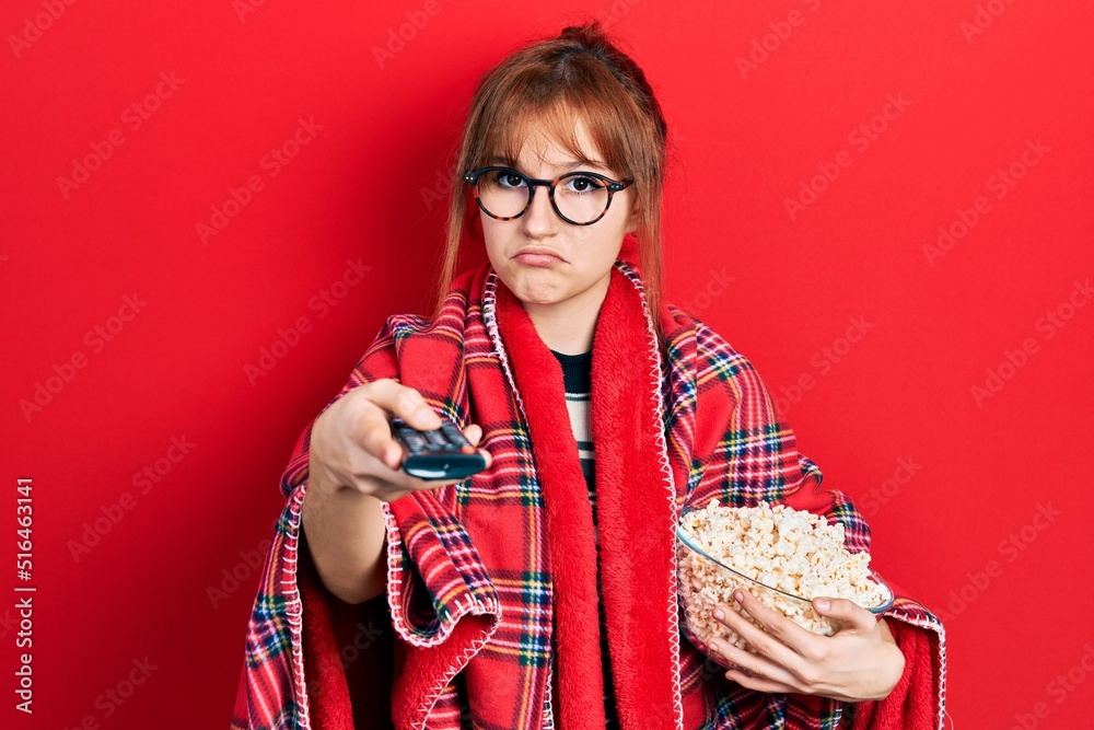 Redhead young woman eating popcorn using tv control depressed and worry ...