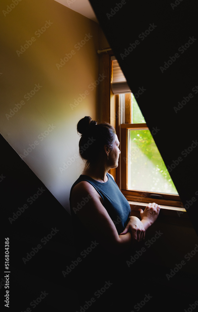 Woman looking out of a dormer window in a dark room in the daytime ...