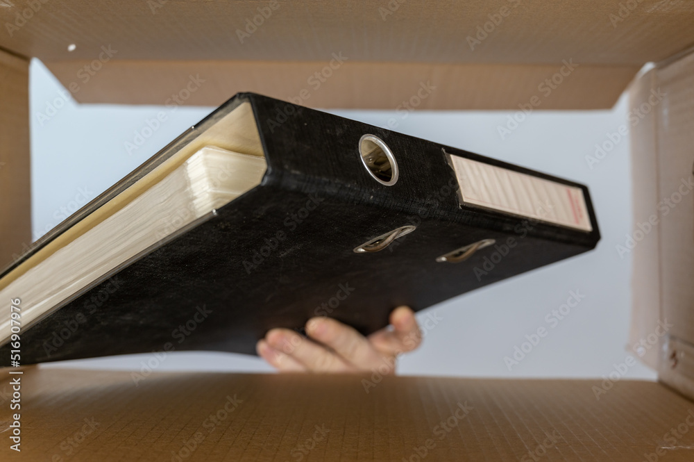 A man's hand places a ring binder folder in a cardboard moving box. A ...