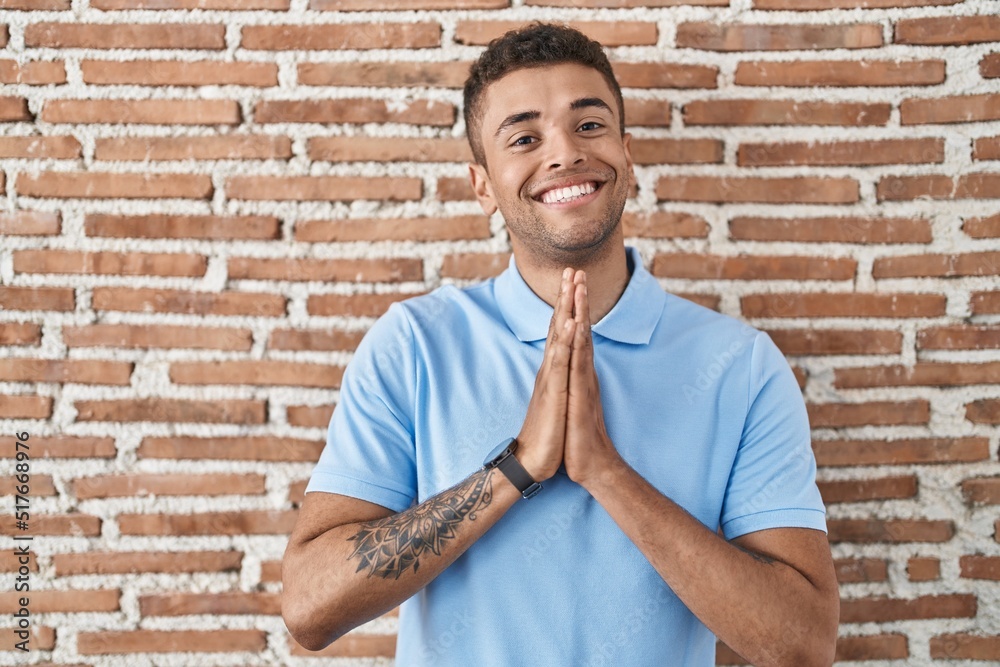 Brazilian young man standing over brick wall praying with hands ...