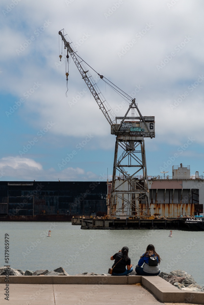 2 sitting people on a curb are looking over water at an old abandoned ...