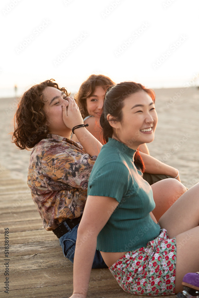3 gen-z friends laughing together while sitting on a beach board walk ...