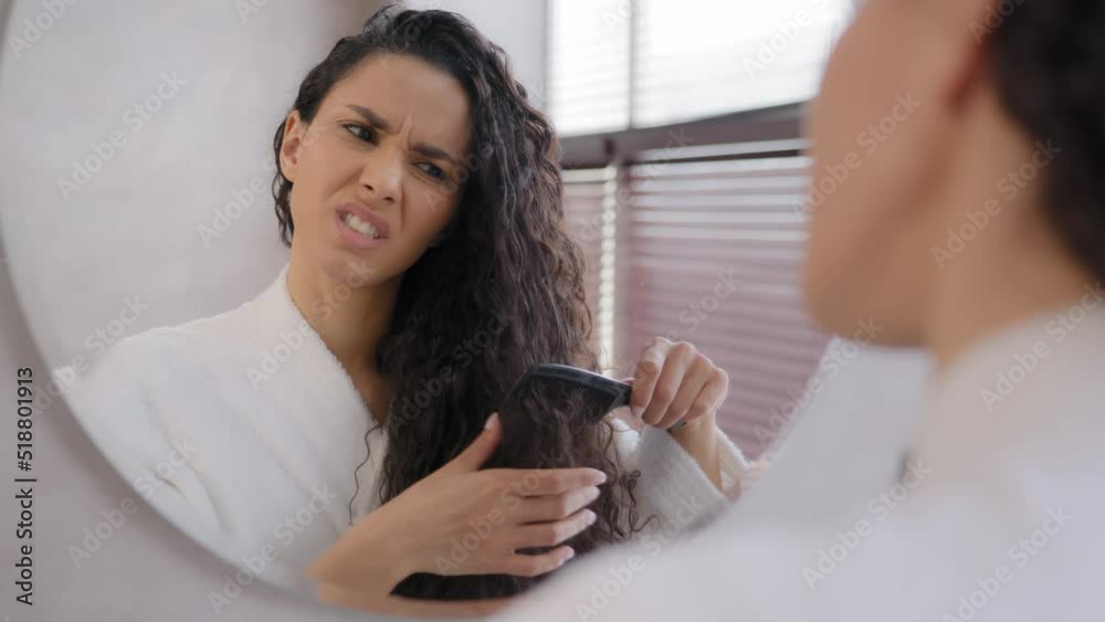 Close-up young woman looking in mirror combing hair in bathroom annoyed ...