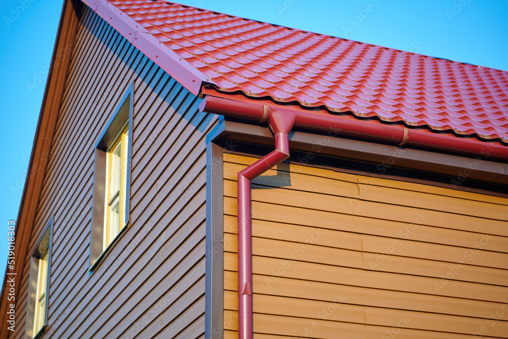 Corner of the house with metal tile roof and rain gutter. Red gutter on ...