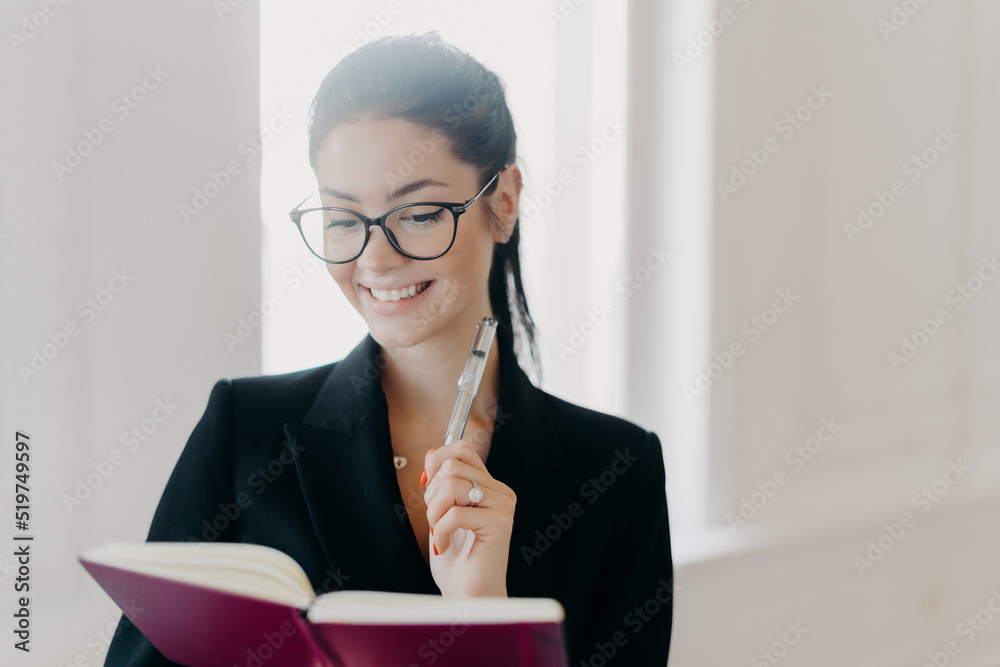 Indoor shot of glad prosperous woman CEO writes in notepad, plans her ...