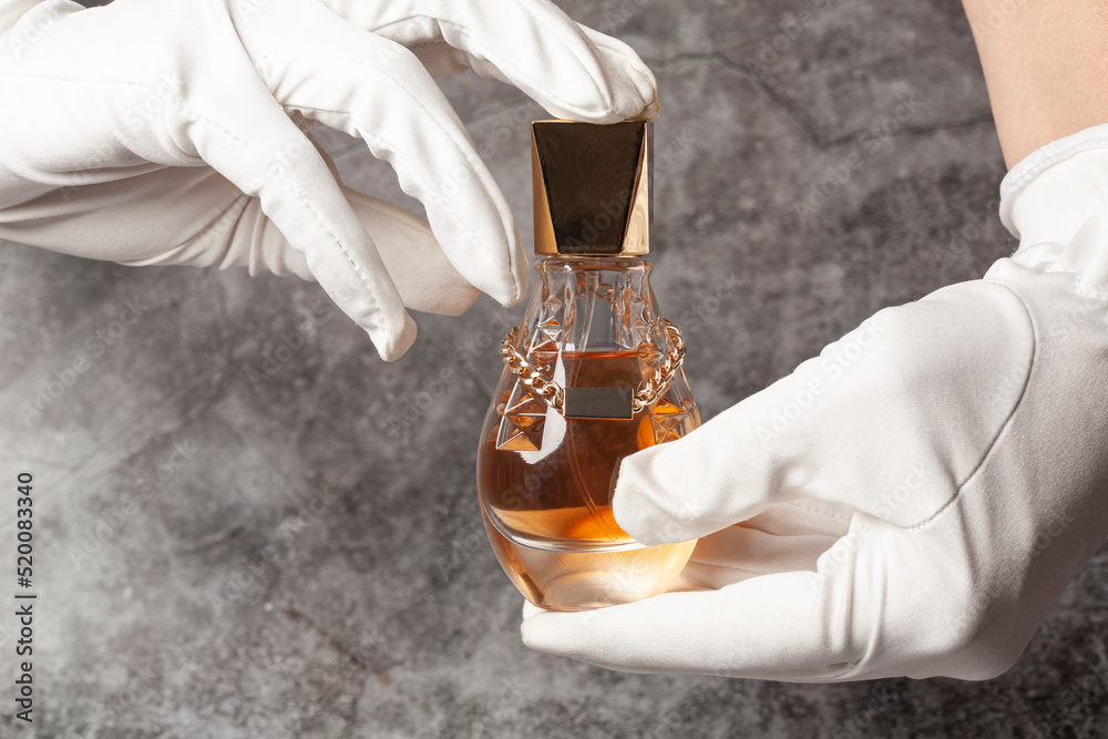 Hands in white gloves holding glass perfume bottle on a gray background ...