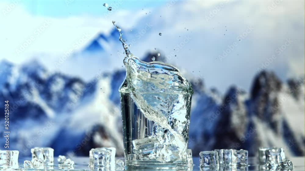 An ice cube falls into a glass of water with splashes. Against the backdrop of mountains. Filmed ...