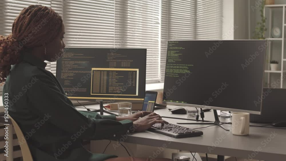 Side view medium slowmo of concentrated young Black woman writing program code on PC sitting in ...