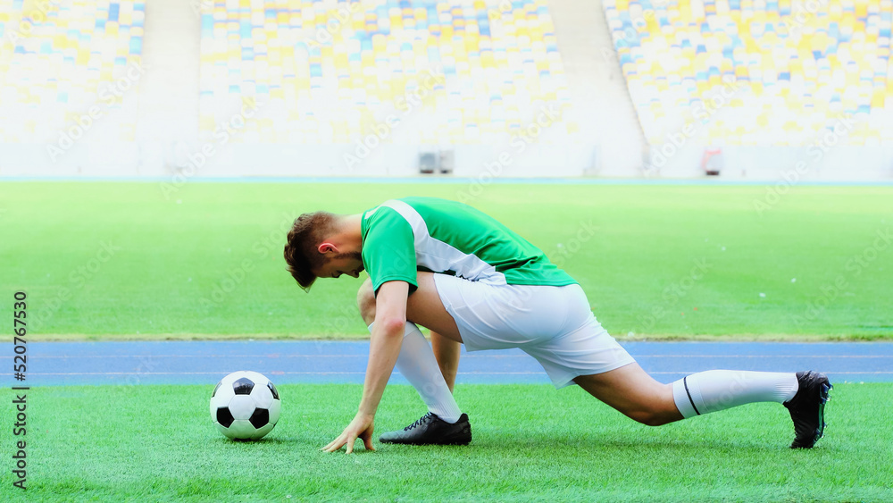 young football player in uniform stretching legs on green grass bear ...