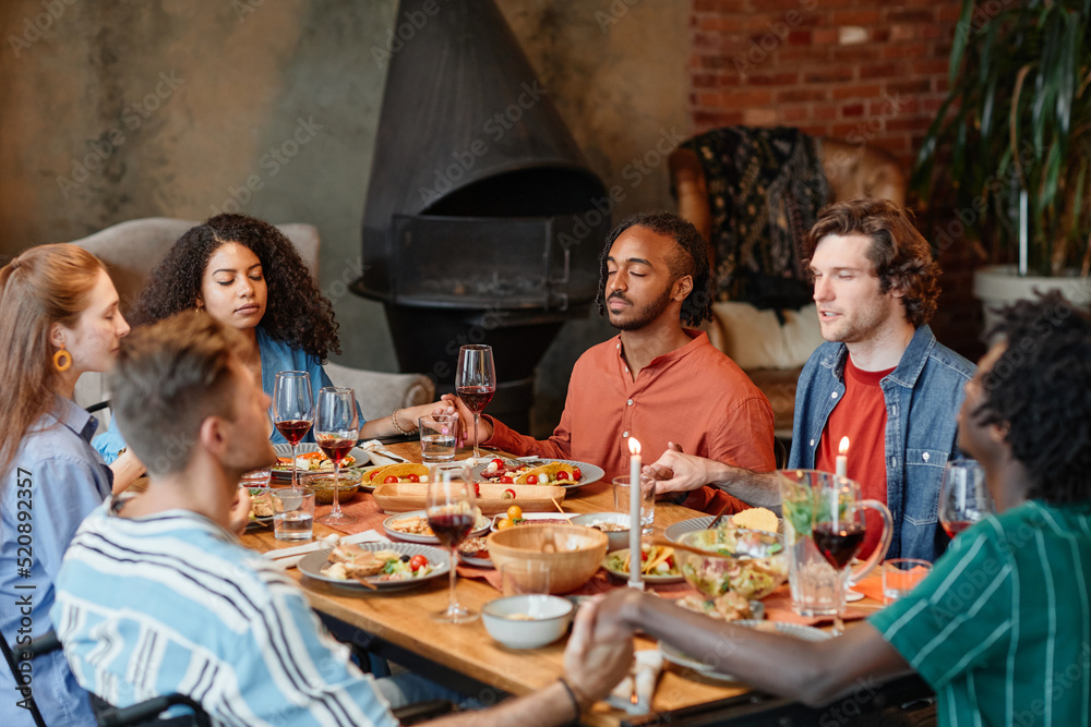 Diverse group of young people holding hands at table and saying grace ...