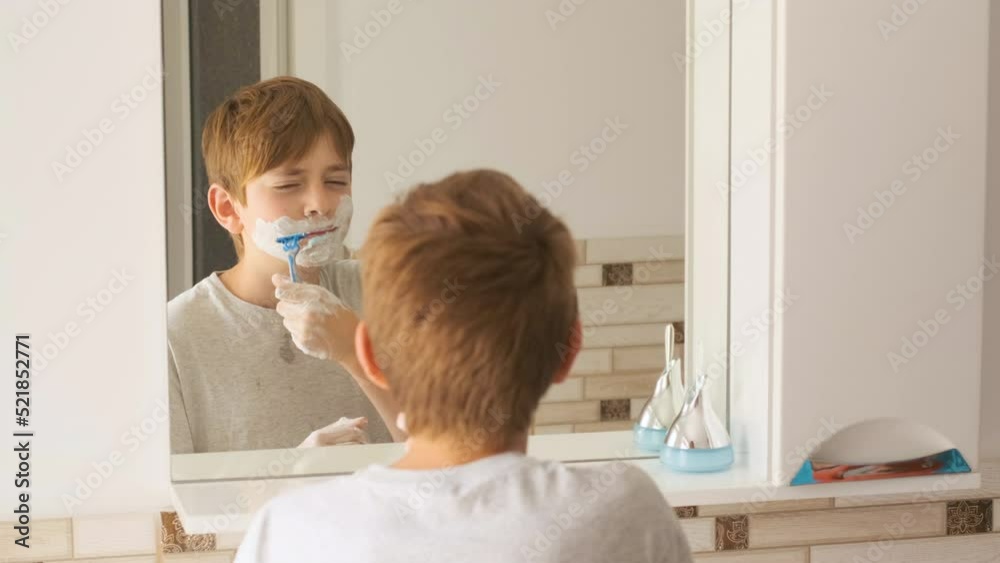 Schoolboy with shaving foam on face, looks carefully in mirror and ...