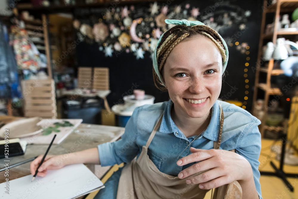 Happy craftsperson holding pencil sitting at workbench in art studio ...