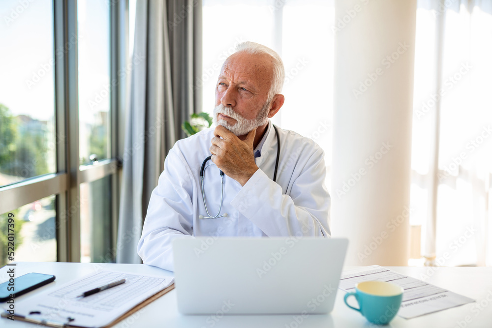Serious mature doctor using laptop and sitting at desk. Senior ...