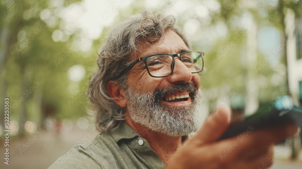 Clouse-up, smiling middle-aged man with gray hair and beard sits on bench and uses mobile phone ...