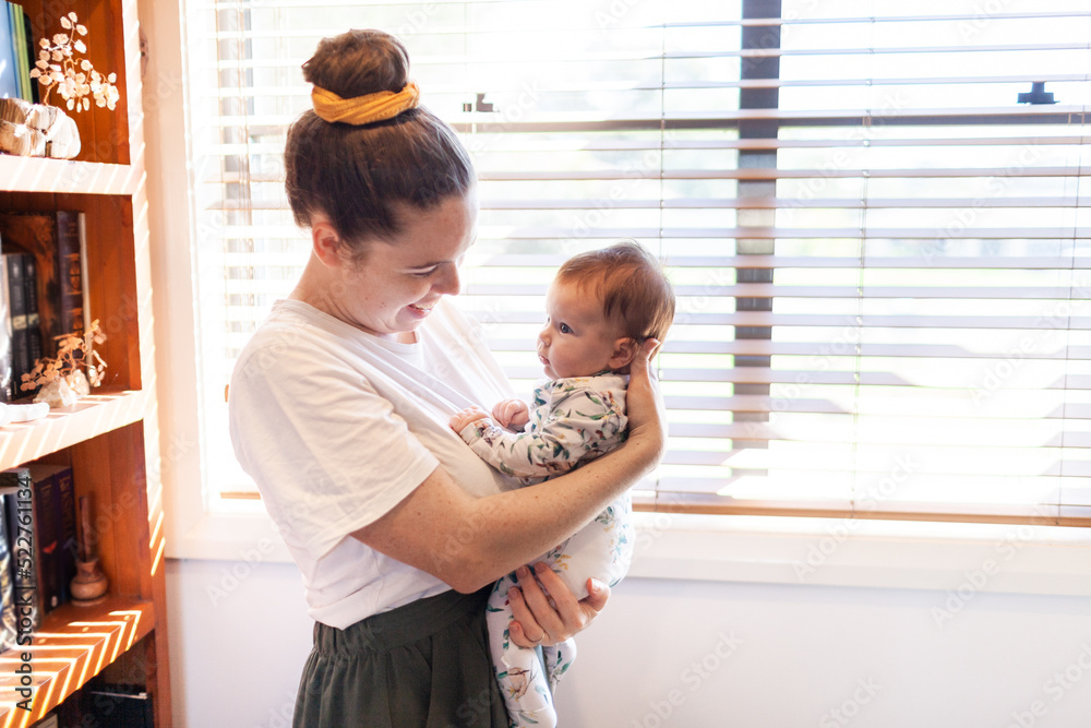 Young smiling mum with hair in a bun holding young baby inside home ...