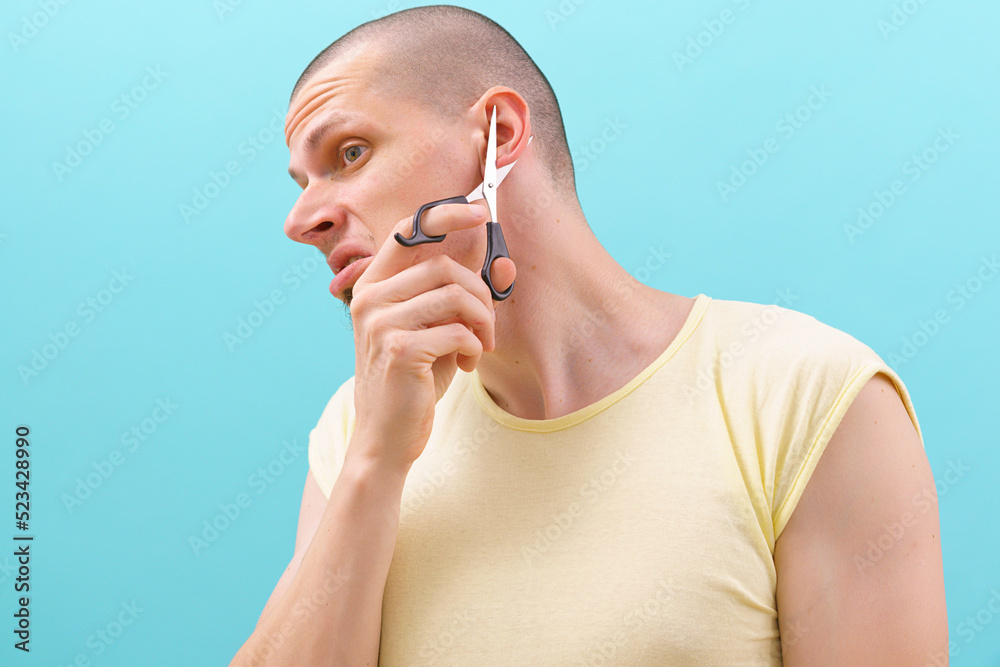 Bald man trying to cut his ear with clippers on a blue background. Aid ...