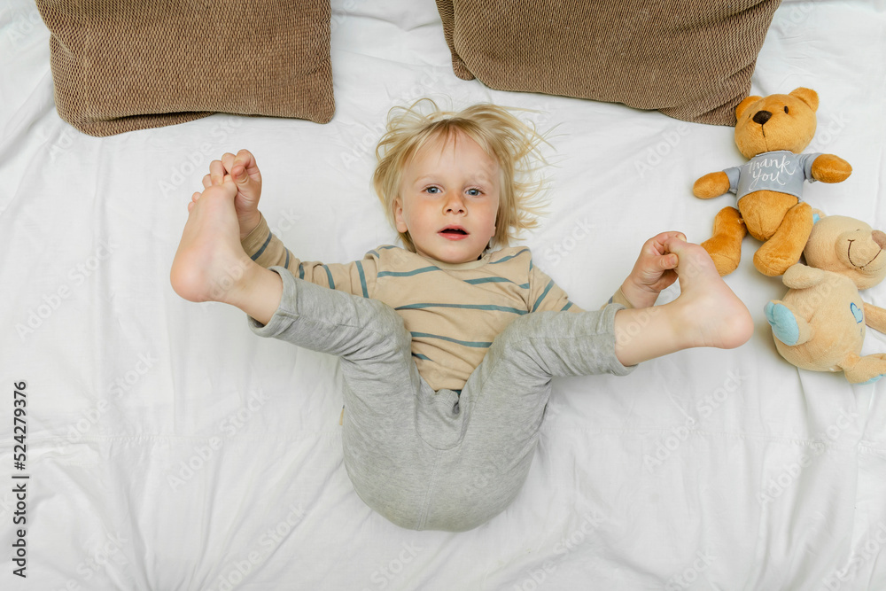 Cute little blond caucasian child kid toddler boy playing,having fun, lying on bed,lifts up the ...