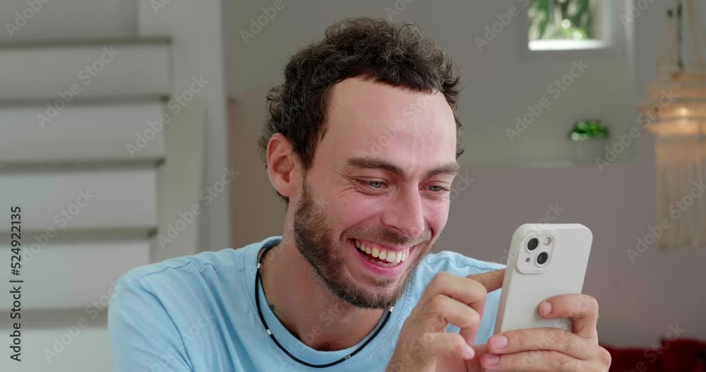 Handsome serene Caucasian young man having fun at home, using his smartphone, scrolling news ...