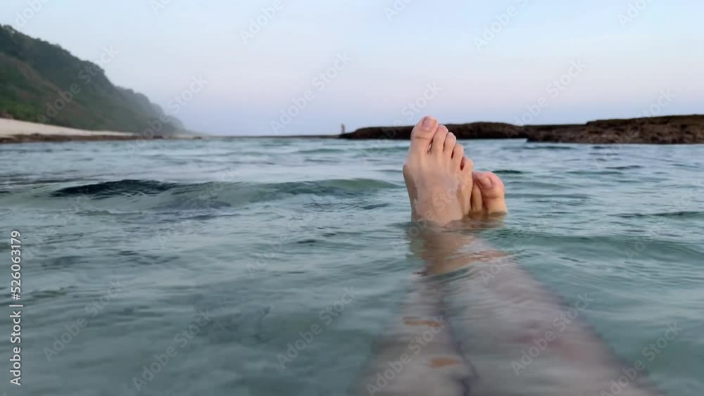 POV view of female feet of woman floating on salty ocean water in tropical paradise. First ...