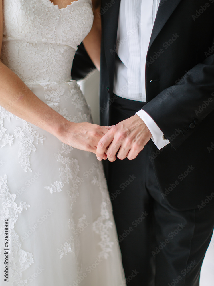 The bride and groom stand hugging and holding hands. The groom is in a ...