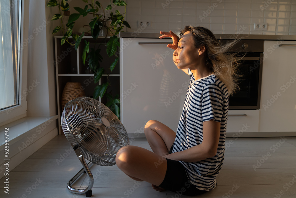 Exhausted young woman refreshing sit near big indoor ventilator blowing ...