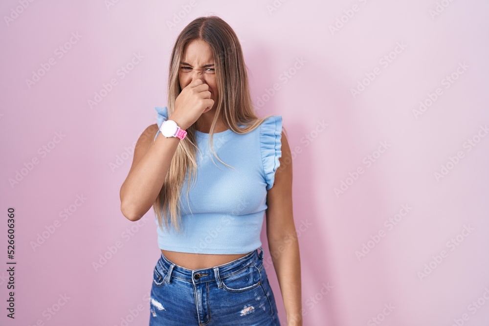 Young blonde woman standing over pink background smelling something ...