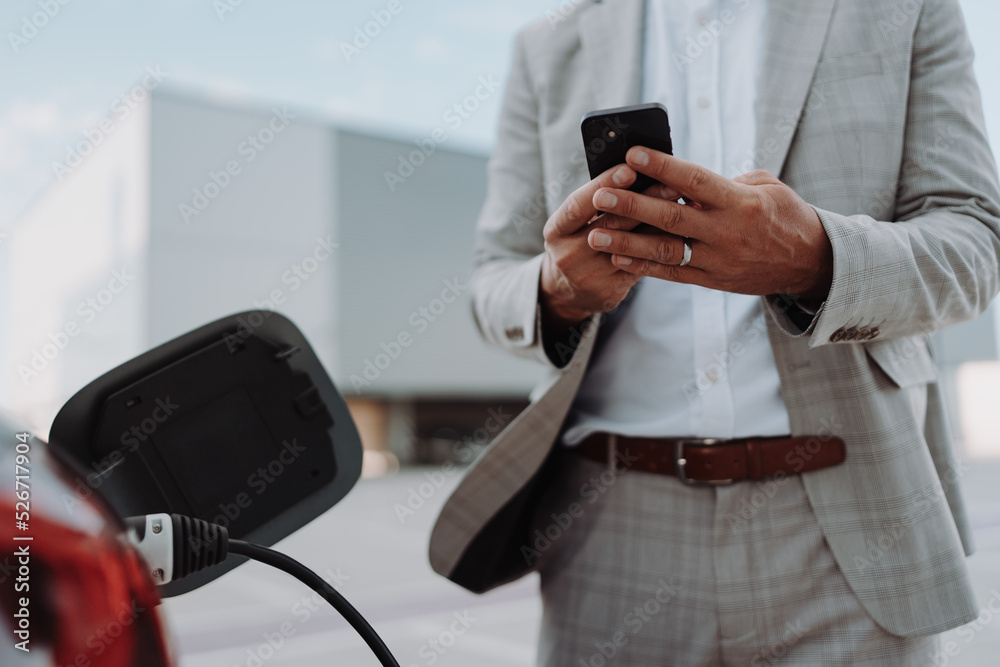 Man holding smartphone while charging car at electric vehicle charging ...