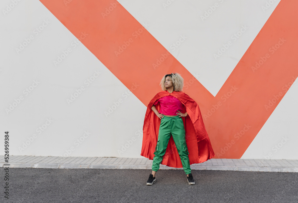 Young woman wearing cape standing with hands on hips in front of wall ...