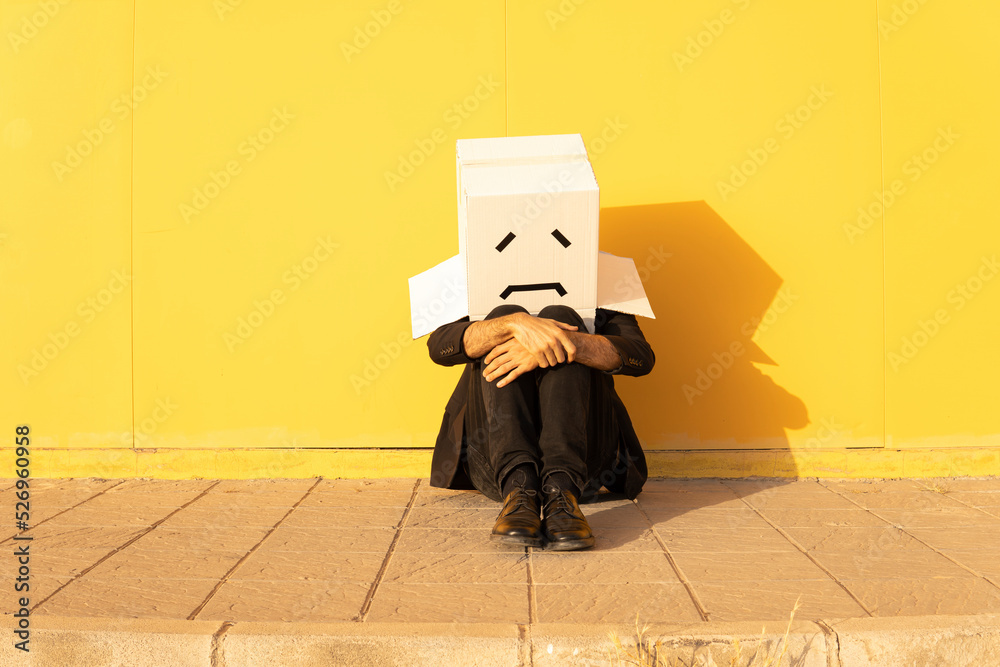 Man wearing box with sad face sitting in front of yellow wall on ...