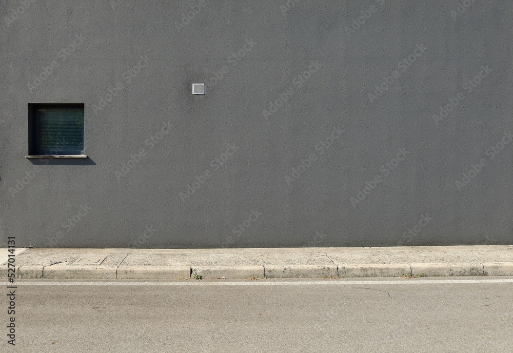 Gray plaster wall with a window and small air inlet. Concrete sidewalk ...