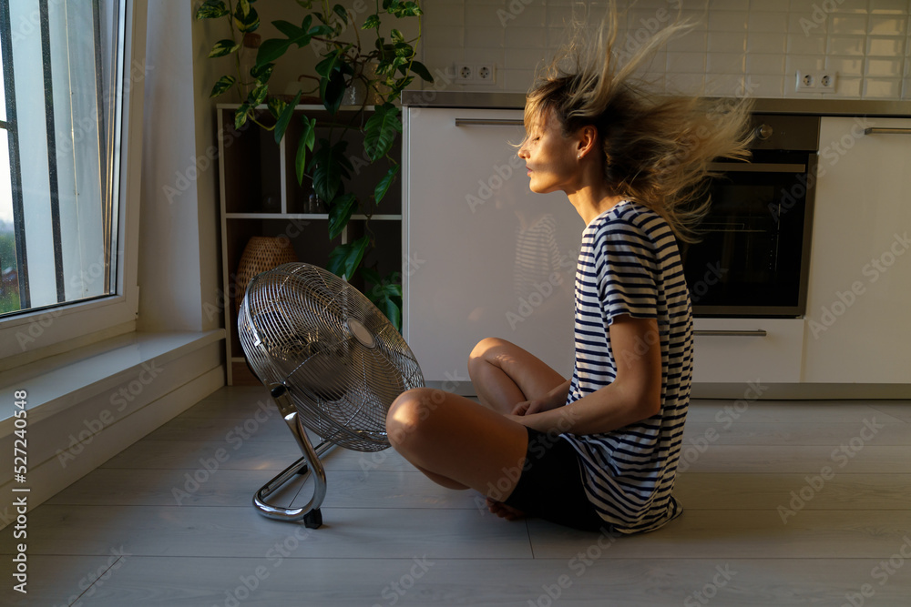 Exhausted young woman refreshing sit near big indoor ventilator blowing ...