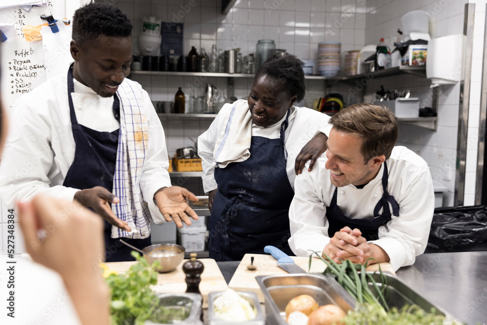 Chef explaining smiling multiracial colleagues at kitchen in restaurant ...