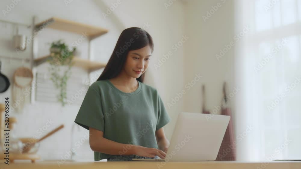 Smiling asian young woman working on laptop at home office. Young asian student using computer ...
