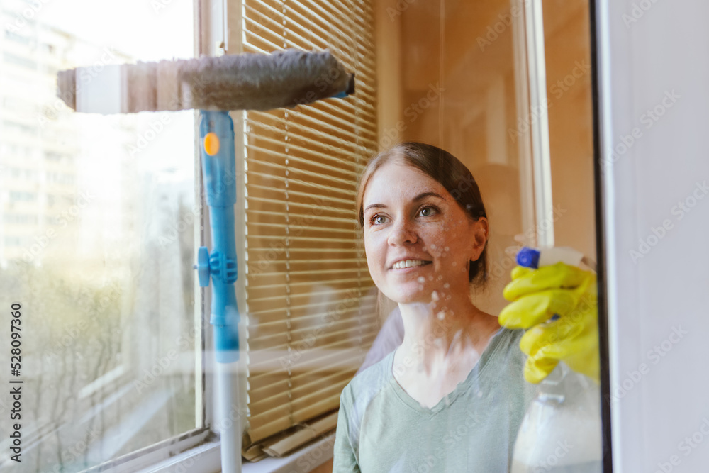 Smiling young woman cleaning window with squeegee and spray detergent ...