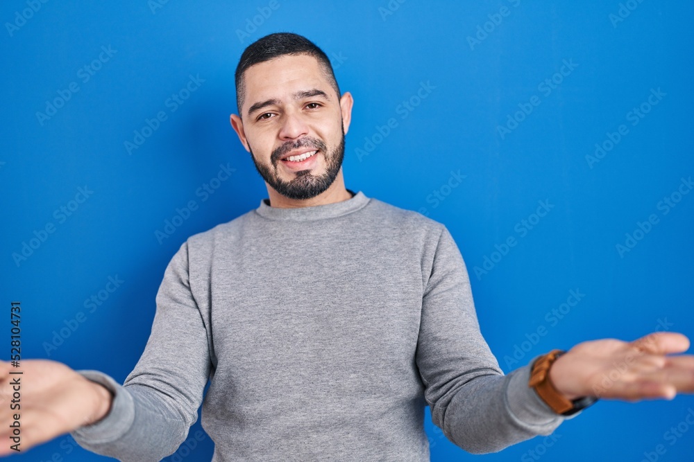 Hispanic man standing over blue background smiling cheerful offering ...